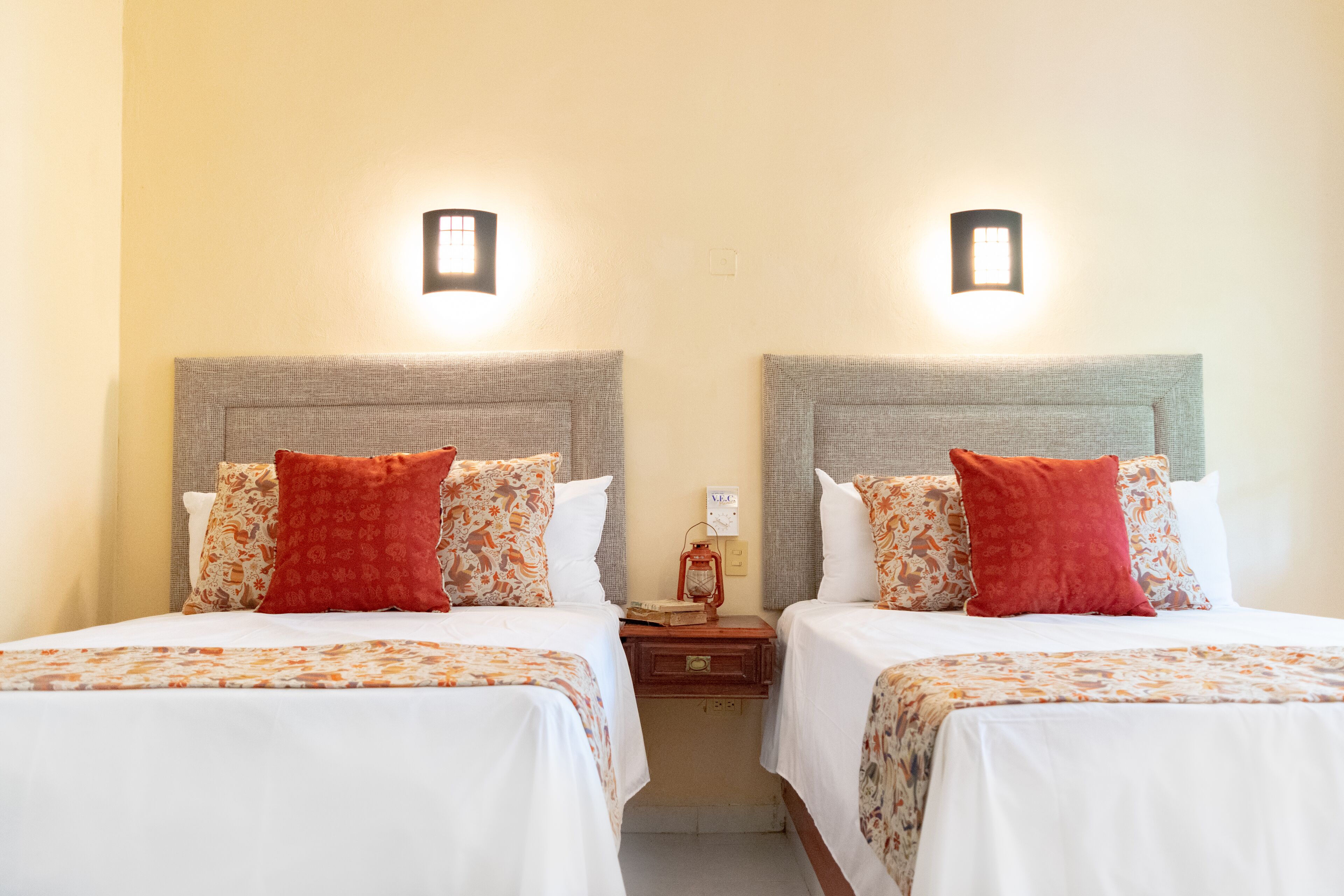 Quarto de hotel com duas camas, cada uma com cabeceira, mesa de cabeceira e luminária fixada na parede.