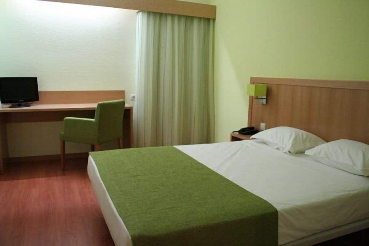 Quarto de hotel com cama, escrivaninha, computador, cadeira e mesa de cabeceira.