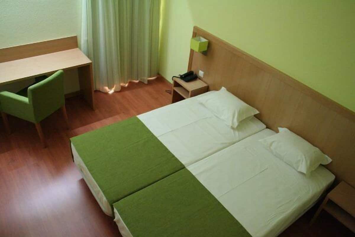 Quarto de hotel com cama, escrivaninha, cadeira e mesa de cabeceira com telefone.