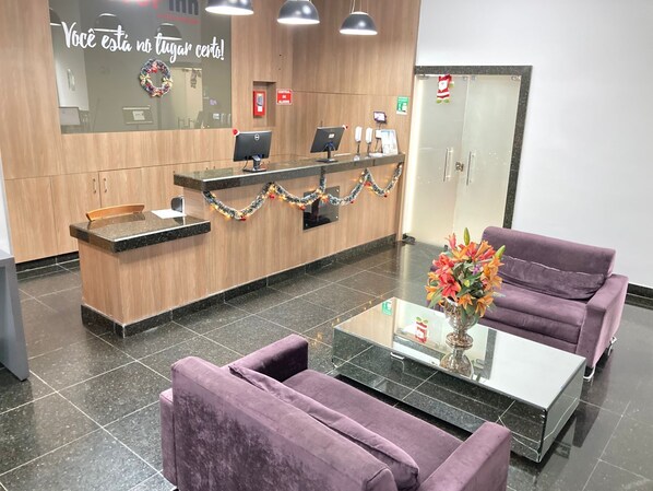 Uma área de recepção com balcão de madeira, dois monitores de computador, um telefone e um guirlanda de Natal. Há dois sofás roxos e uma mesa com um vaso de flores.
