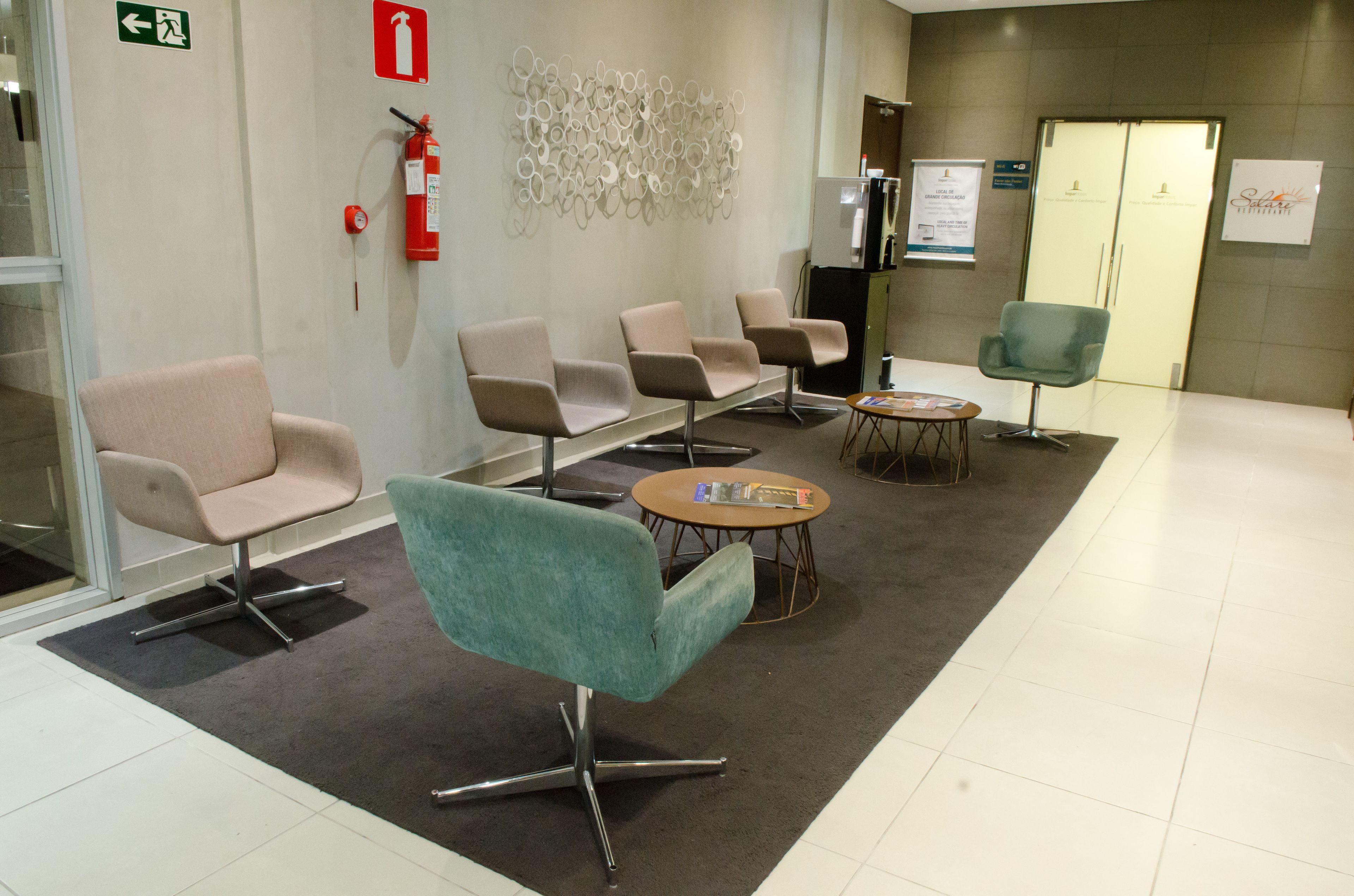 Uma área de espera com várias cadeiras, uma pequena mesa, um extintor de incêndio e um objeto decorativo fixado na parede.