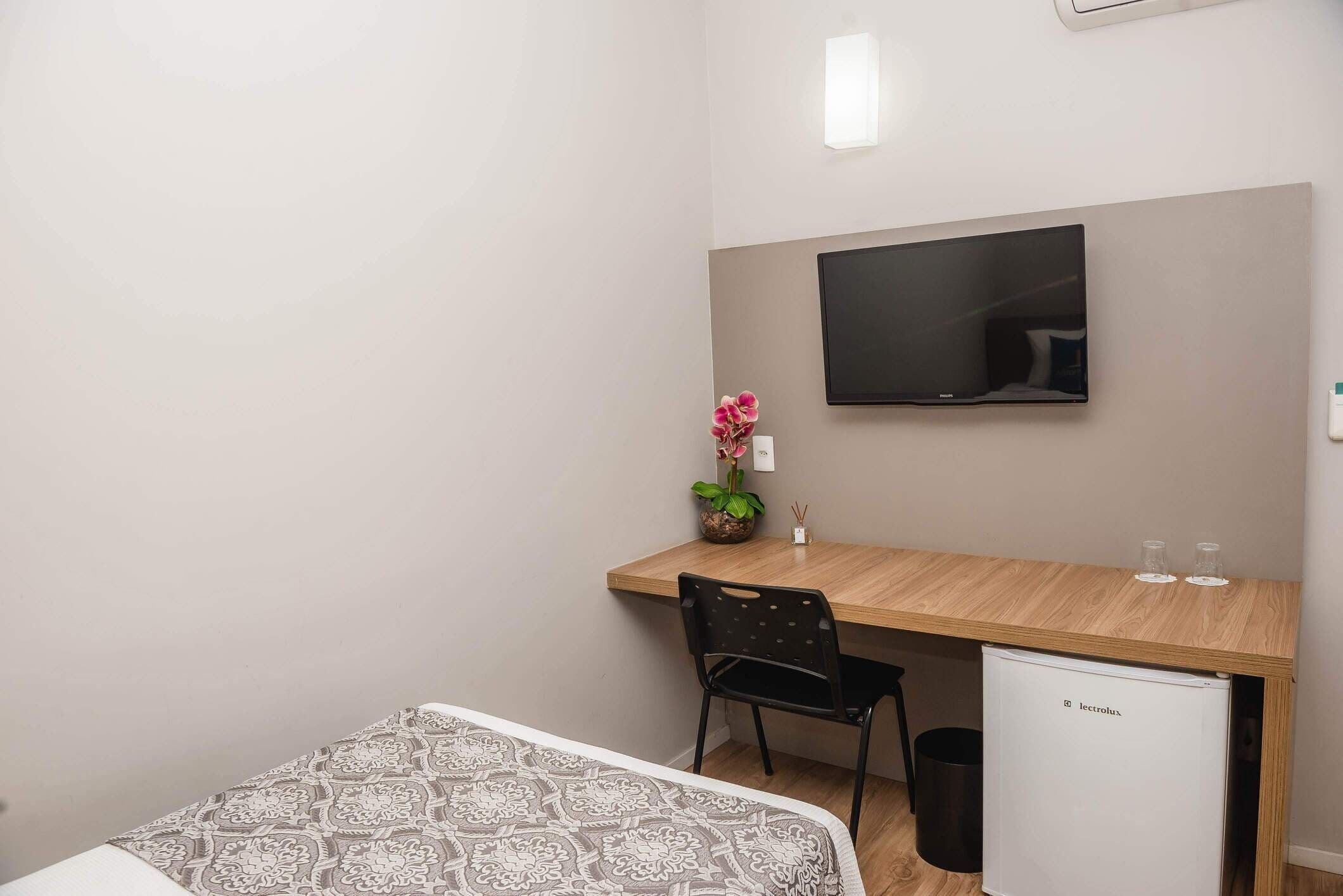 Quarto de hotel compacto com uma cama, uma escrivaninha com cadeira, televisão, mini-geladeira e lixeira.