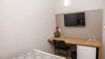 Quarto de hotel compacto com uma cama, uma escrivaninha com cadeira, televisão, mini-geladeira e lixeira.