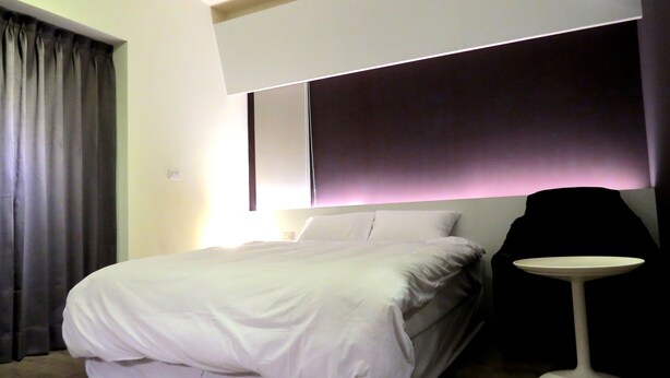 Quarto de hotel com cama, mesa de cabeceira, televisão na parede e janela com cortinas.