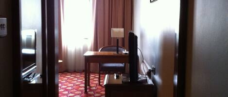 Quarto de hotel com carpete vermelho estampado, uma escrivaninha com cadeira, televisão e janela com cortinas.