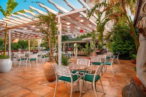 Um pátio externo com mobiliário de vime branco e almofadas verdes, cercado por plantas em vasos e uma estrutura de pérgola.