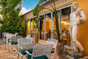 Um pátio externo com cadeiras brancas ricamente decoradas, uma mesa e uma estátua de uma figura clássica.