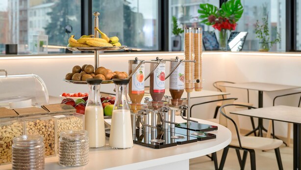 Uma mesa de pequeno-almoço com diversos alimentos, incluindo frutas, frutos secos e bebidas, e vista para a cidade através de grandes janelas.