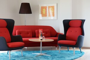 Quarto moderno com sofá vermelho, duas poltronas preto e vermelho, uma mesa redonda pequena com uma garrafa e um copo, e um tapete azul.