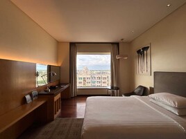 Quarto de hotel com cama, escrivaninha, cadeira, televisão e uma grande janela com vista para a cidade.