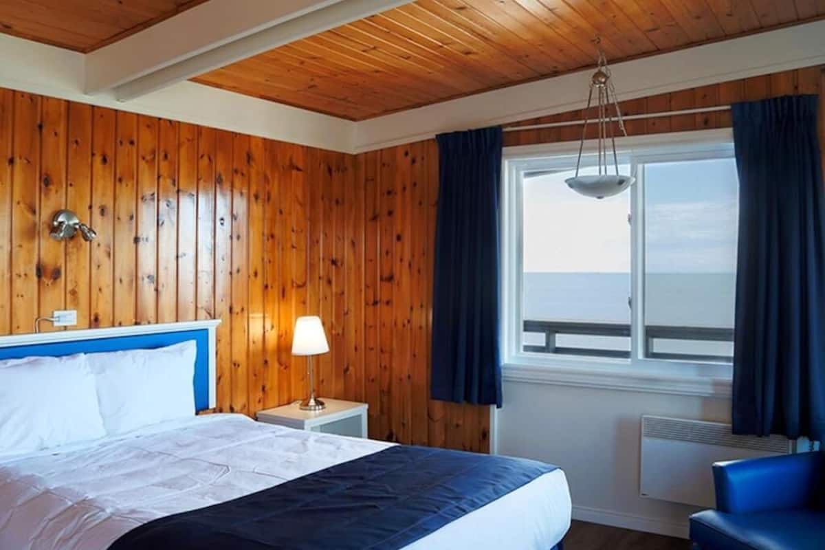 Quarto com parede de painel de madeira, cama com roupa de cama branca e azul, mesa de cabeceira com abajur, janela com cortinas e vista para o mar.