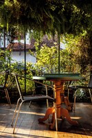 Um pátio externo com mesa e cadeiras, cercado por vegetação exuberante e vista para um edifício com telhado de azulejos.