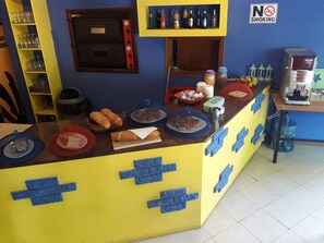 Uma área de cozinha com bancada exibindo diversos alimentos, uma máquina de café e um aviso de 'Não Fumar' na parede.