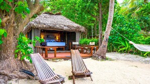 Uma cabana com telhado de palha, com cama e área de refeições, cercada por vegetação exuberante e uma rede.