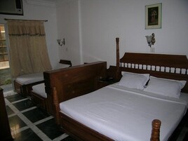 Quarto com uma cama de madeira grande, uma cama menor, uma mesinha de cabeceira, um abajur e um quadro na parede.
