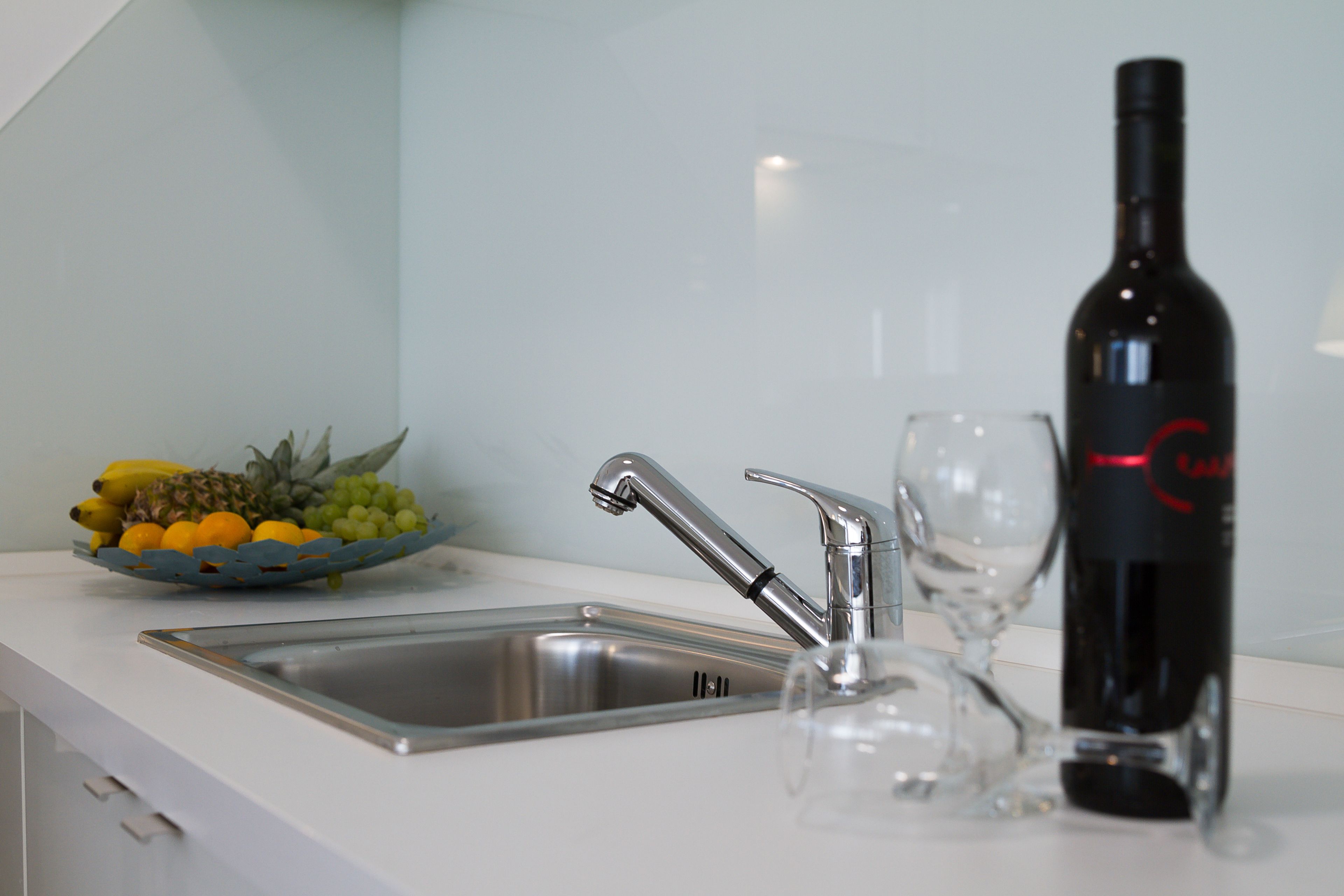 Uma bancada de cozinha com uma pia de aço inoxidável, uma tigela de frutas, uma garrafa de vinho e uma taça de vinho.