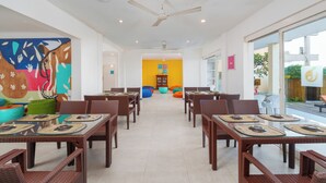Uma área de jantar com móveis de vime, um mural colorido e uma parede amarelo vibrante ao fundo.