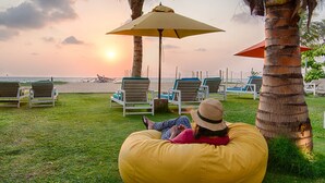 Uma área de descanso à beira-mar com cadeiras e um grande guarda-sol, uma pessoa relaxando em uma poltrona de espuma, e um pôr do sol ao fundo.