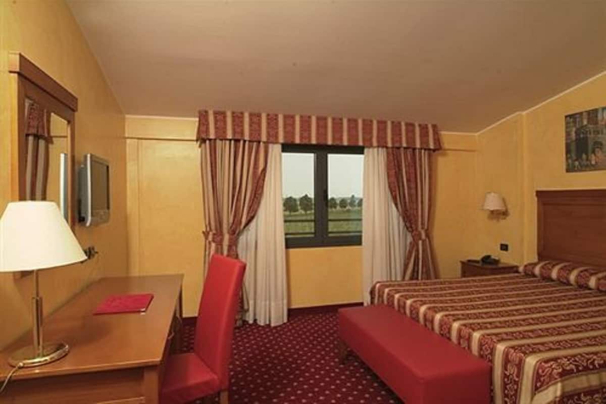 Quarto de hotel com cama, escrivaninha, cadeira, luminária e janela com cortinas.