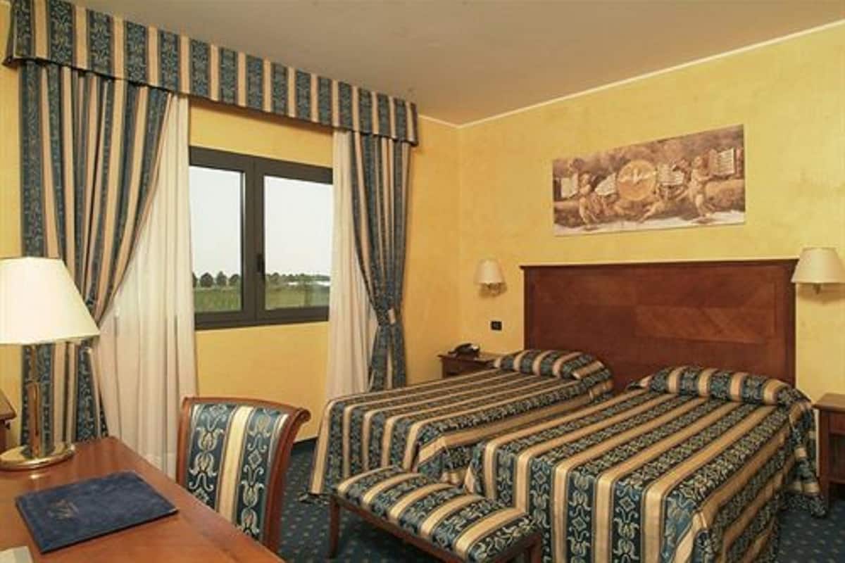 Quarto de hotel com duas camas, uma cabeceira de madeira, uma mesa de jantar e uma janela com cortinas.