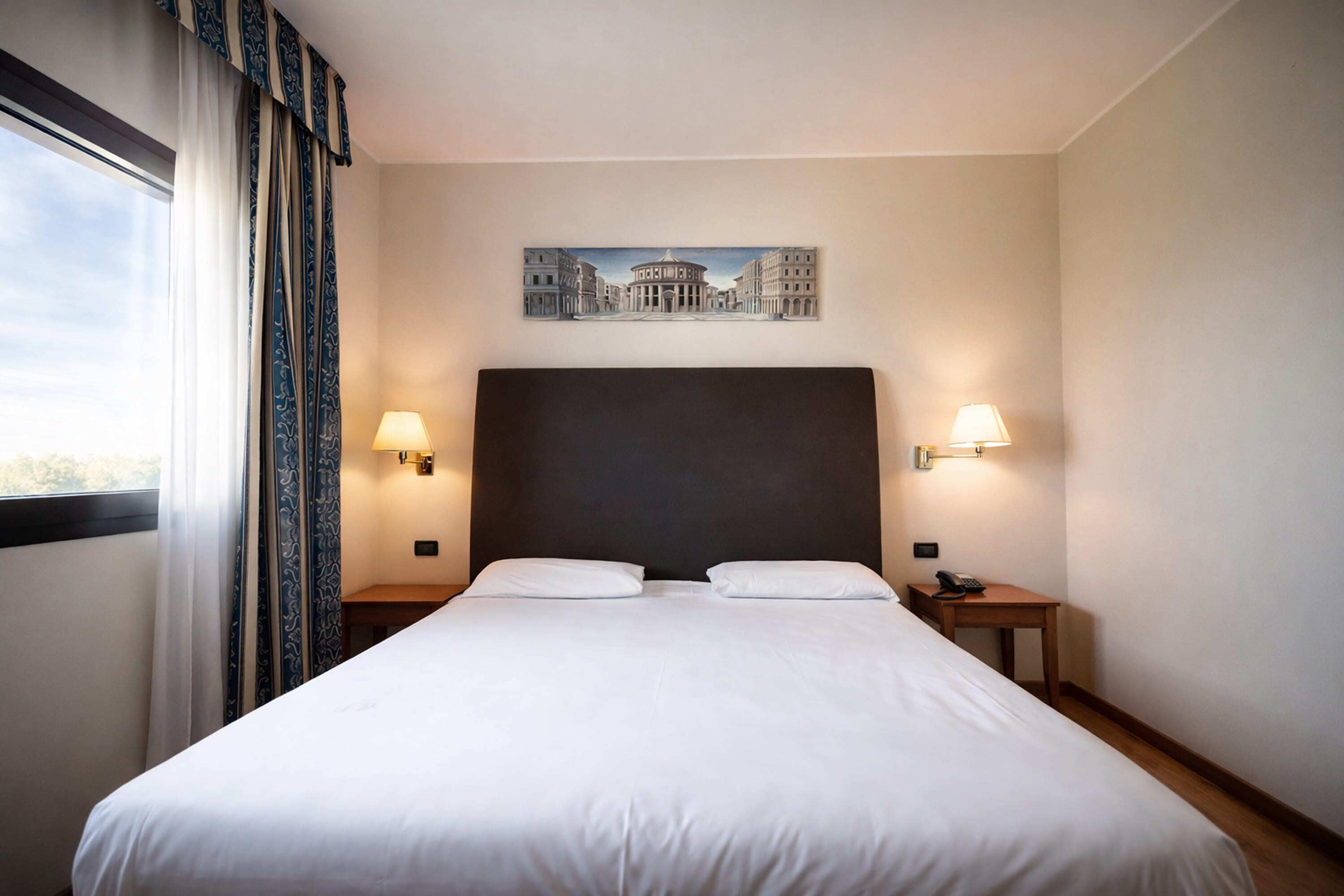Quarto de hotel com uma cama grande, duas luminárias de cabeceira, um quadro de um edifício na parede e uma janela com cortinas.