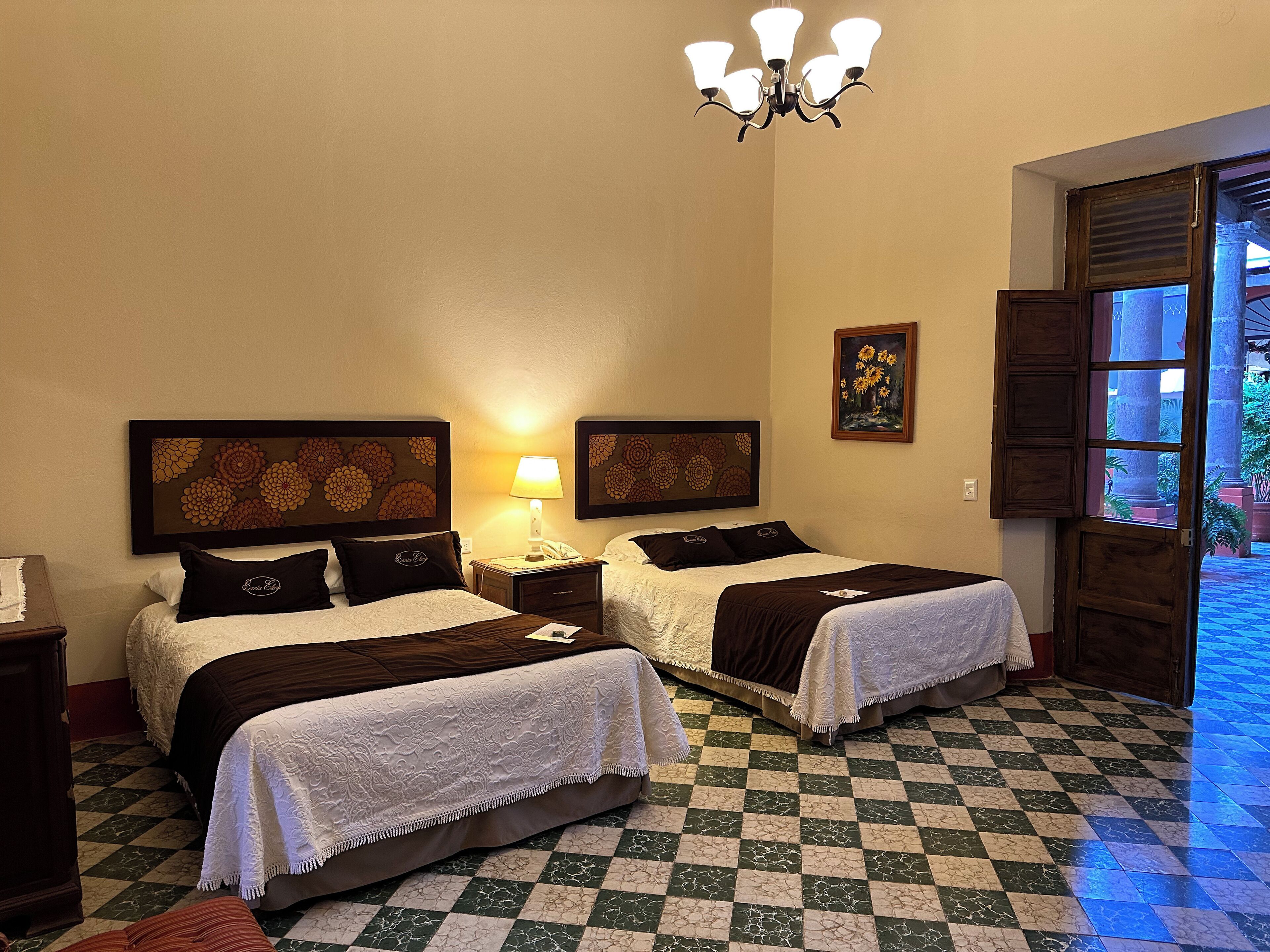 Quarto de hotel com duas camas, um candelabro, obras de arte nas paredes e chão xadrez.