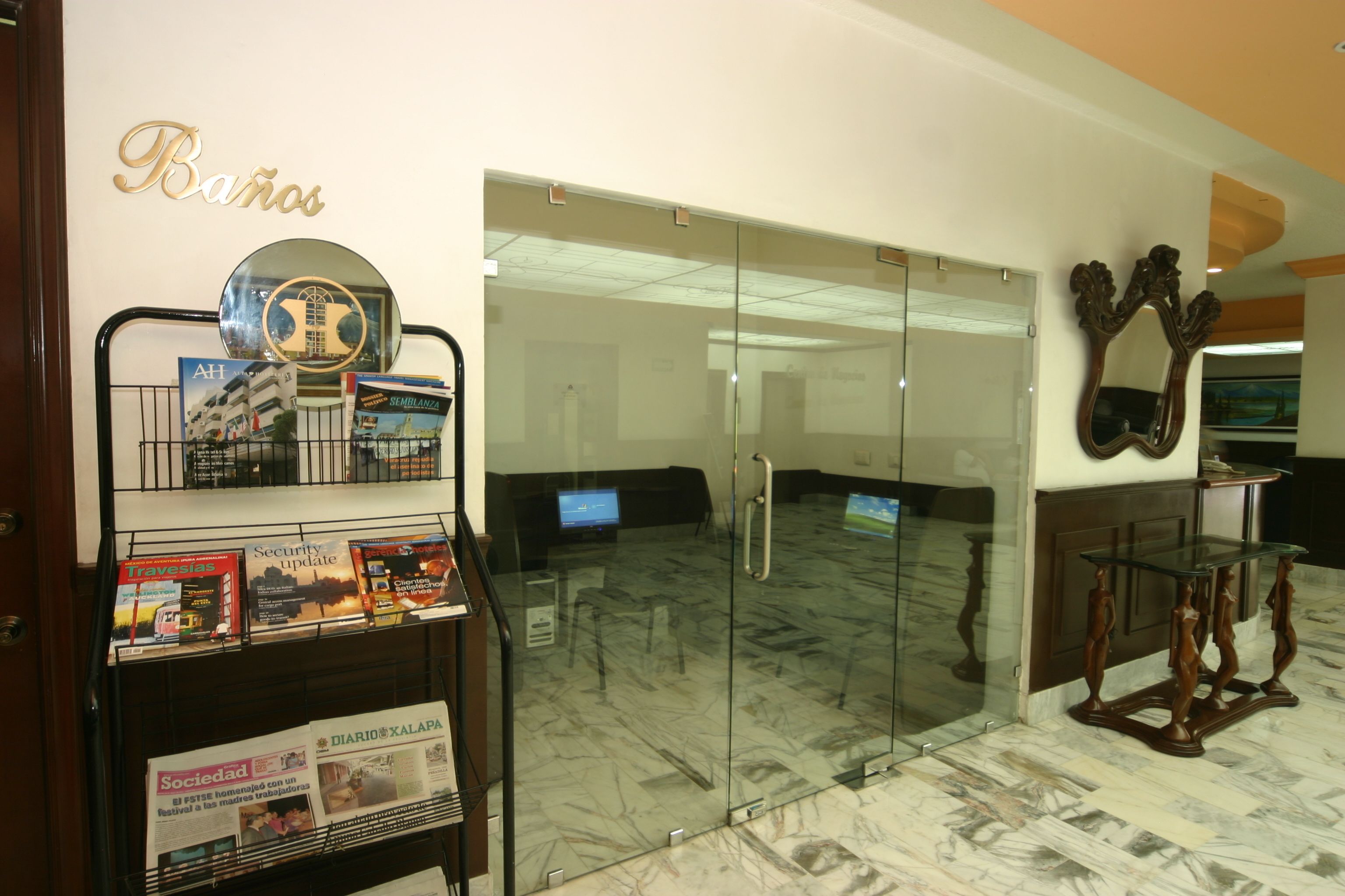 Uma área de recepção com porta de vidro, espelho fixado na parede e mesa de madeira com revistas.