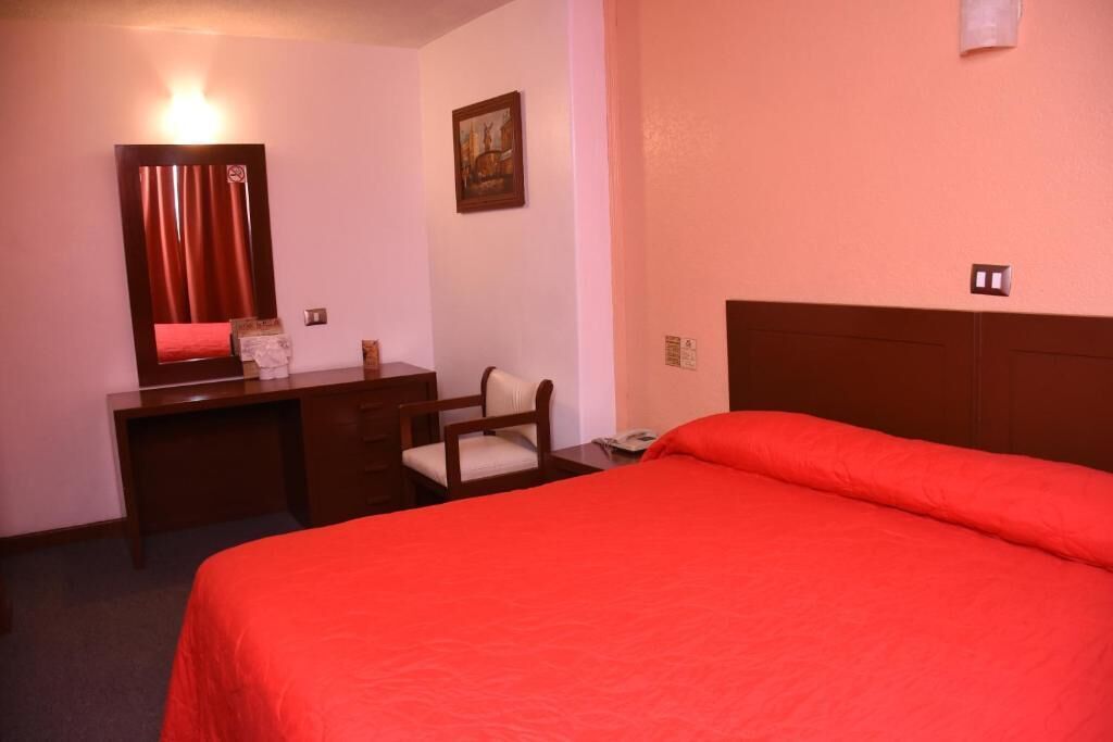 Quarto de hotel com uma cama grande, uma escrivaninha com cadeira, um espelho e uma luminária fixada na parede.