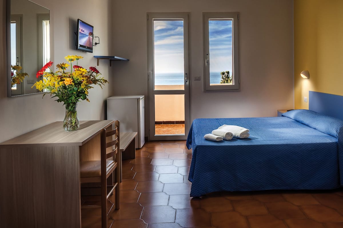 Quarto com cama, escrivaninha com cadeira, vaso com flores, televisão e vista para o mar através de uma janela.