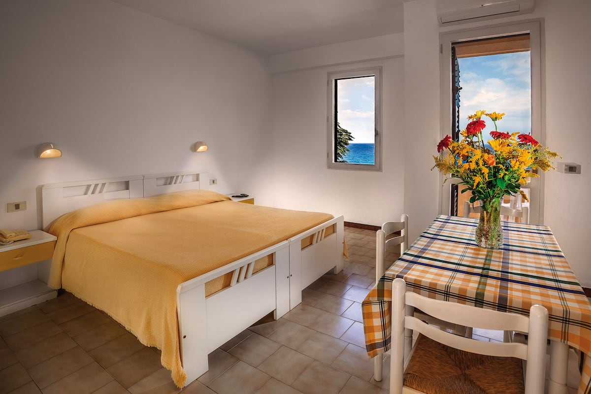 Quarto com cama, mesa de cabeceira e janela com vista para o mar.