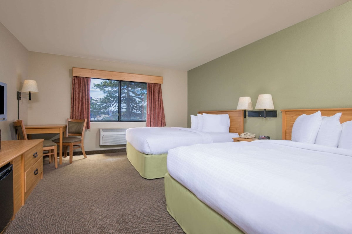 Quarto de hotel com duas camas, uma escrivaninha, uma cadeira, um abajur e uma janela com cortinas.