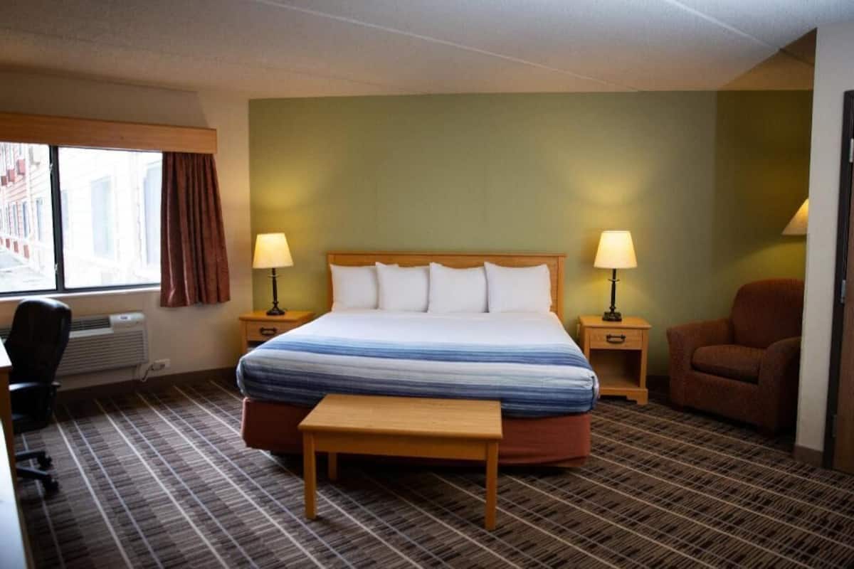 Quarto de hotel com uma cama grande, duas mesas de cabeceira com luminárias, um banco de madeira, uma escrivaninha, uma cadeira e uma janela com cortinas.