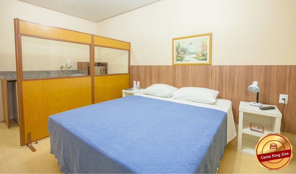 Quarto de hotel com cama, mesa de cabeceira, escrivaninha com luminária, um quadro na parede e guarda-roupa com espelho.