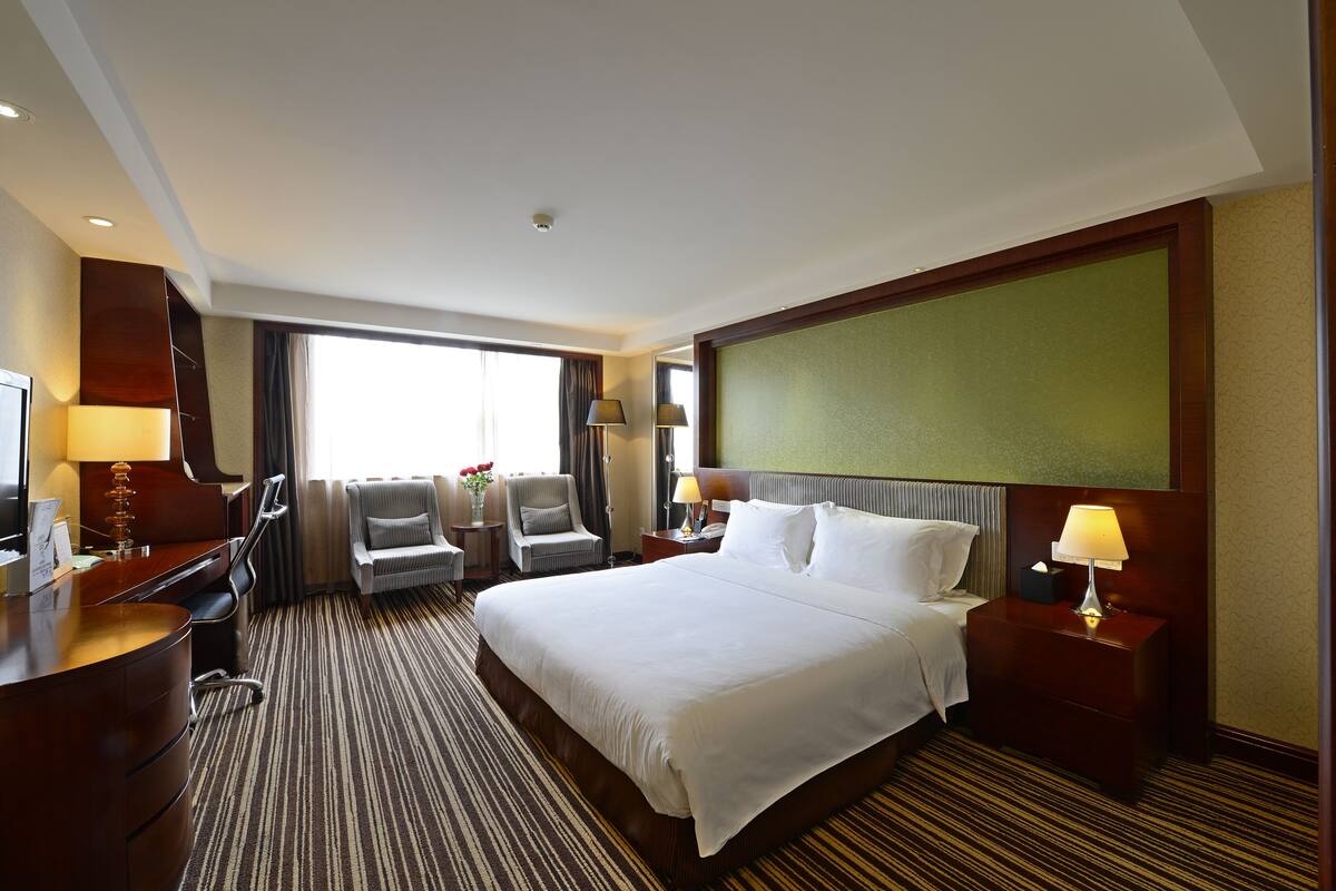Quarto de hotel com uma cama grande, duas poltronas, uma escrivaninha com abajur e uma televisão.