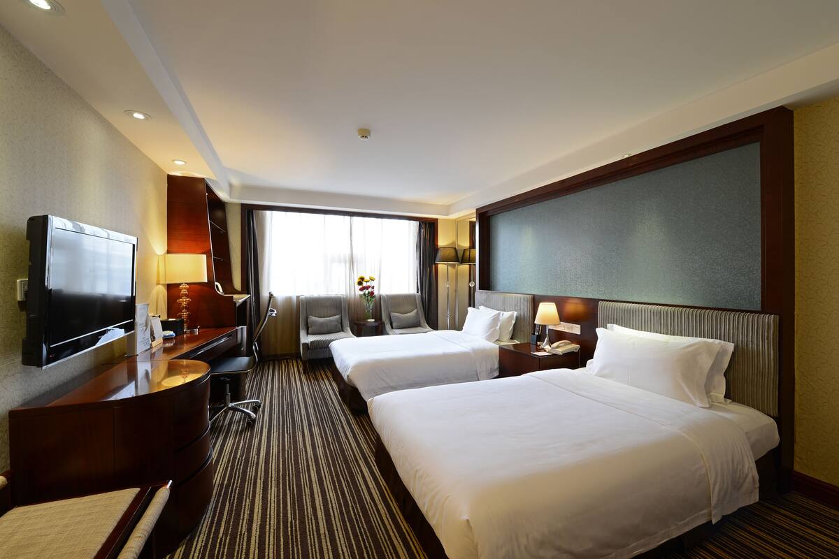 Quarto de hotel com duas camas, uma escrivaninha com televisão, uma cadeira e uma janela grande com cortinas.