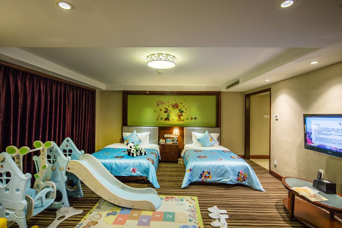 Quarto de hotel com duas camas, televisão, escrivaninha, cadeira, mesa pequena, luminária, cortina, decoração na parede e um escorrega.