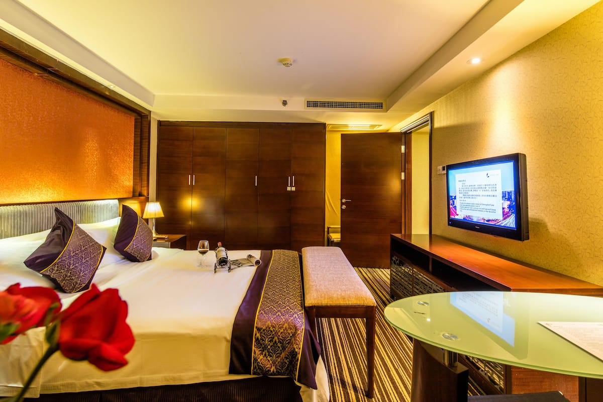Quarto de hotel com uma cama, uma escrivaninha, uma televisão e uma mesa de vidro.
