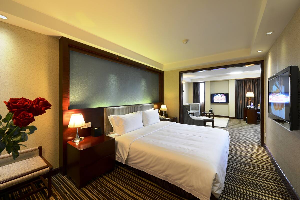 Quarto de hotel com uma cama grande, mesa de cabeceira com abajur, uma cadeira, escrivaninha com televisão e vista para o corredor.
