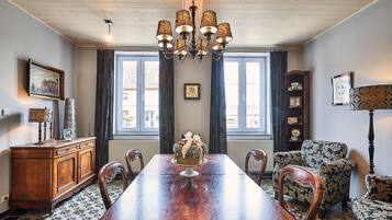 Uma sala de jantar com mesa de madeira, cadeiras, um lustre e vista através de grandes janelas.