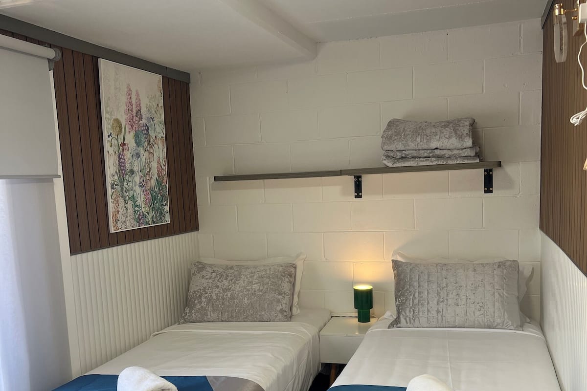Quarto de hotel com duas camas, uma prateleira na parede com uma toalha, abajur e ventilador de teto.