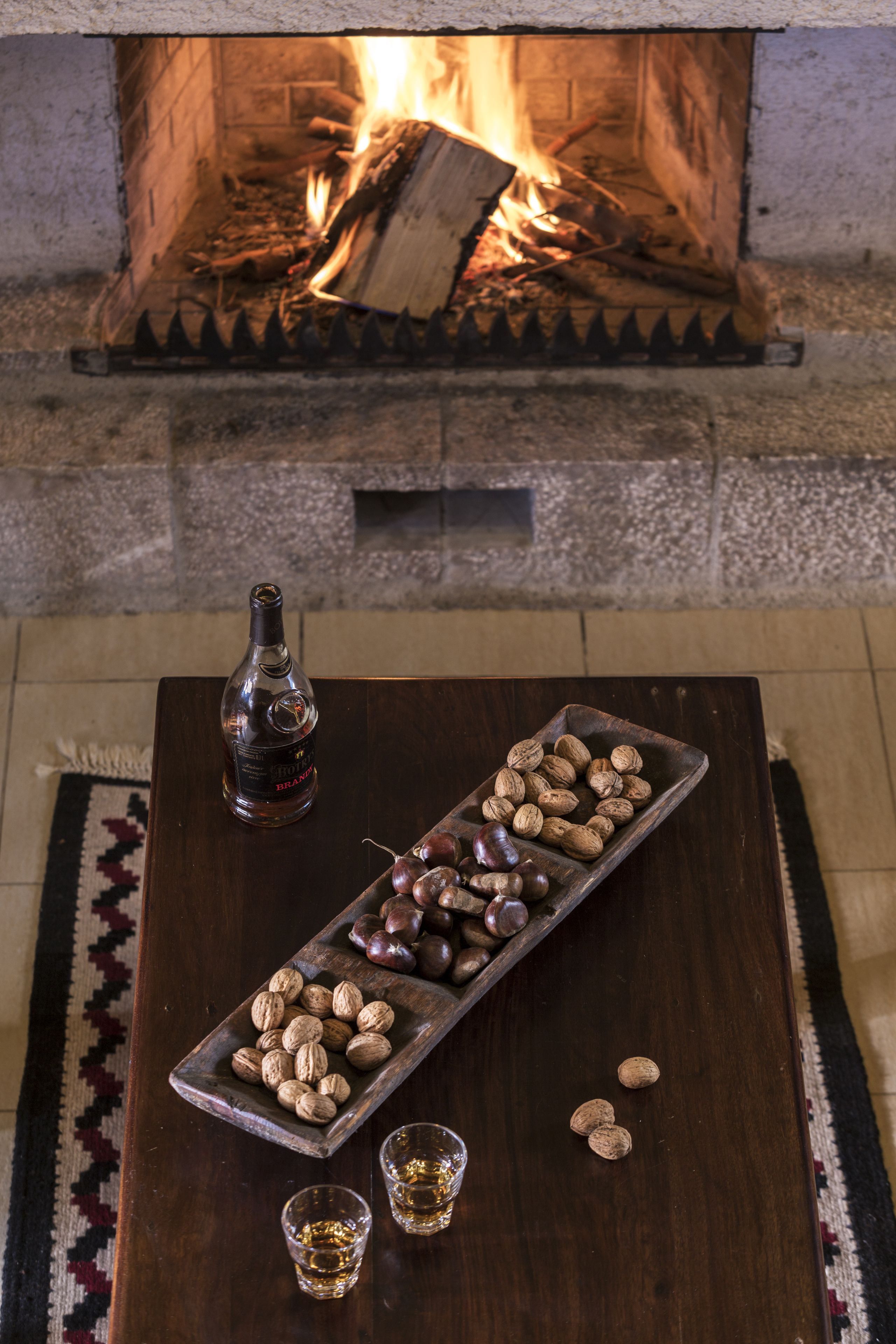 Uma mesa de madeira com uma garrafa e copos, uma bandeja de nozes e uma lareira ao fundo.