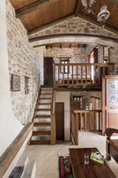 Interior rústico com parede de pedra, escada de madeira e um quarto no mezanino com cama e janela.