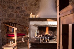 Quarto rústico com parede de pedra, uma cadeira de madeira com almofada vermelha, uma mesa de madeira com uma garrafa, copos e uma tigela de nozes, e uma lareira com velas acesas.