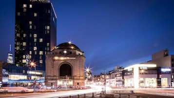 Um edifício histórico com uma grande entrada em arco, rodeado por edifícios modernos e ruas movimentadas à noite.