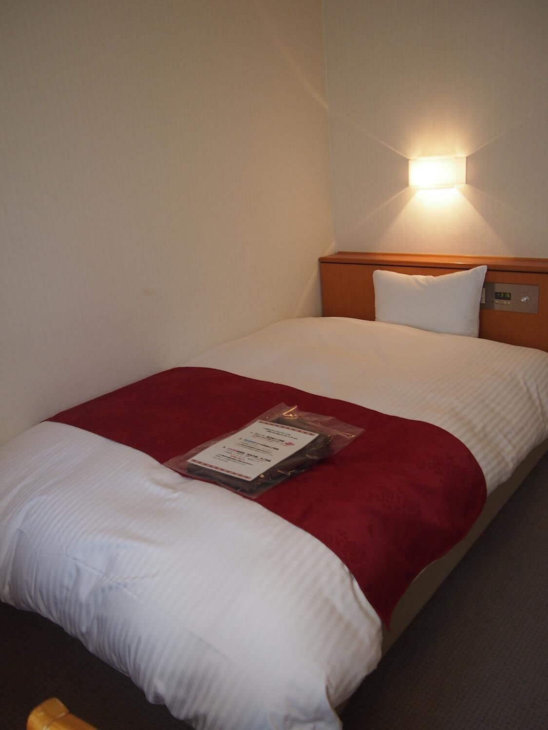 Quarto de hotel com uma cama bem arrumada, um cobertor vermelho e uma mesa de cabeceira com abajur.