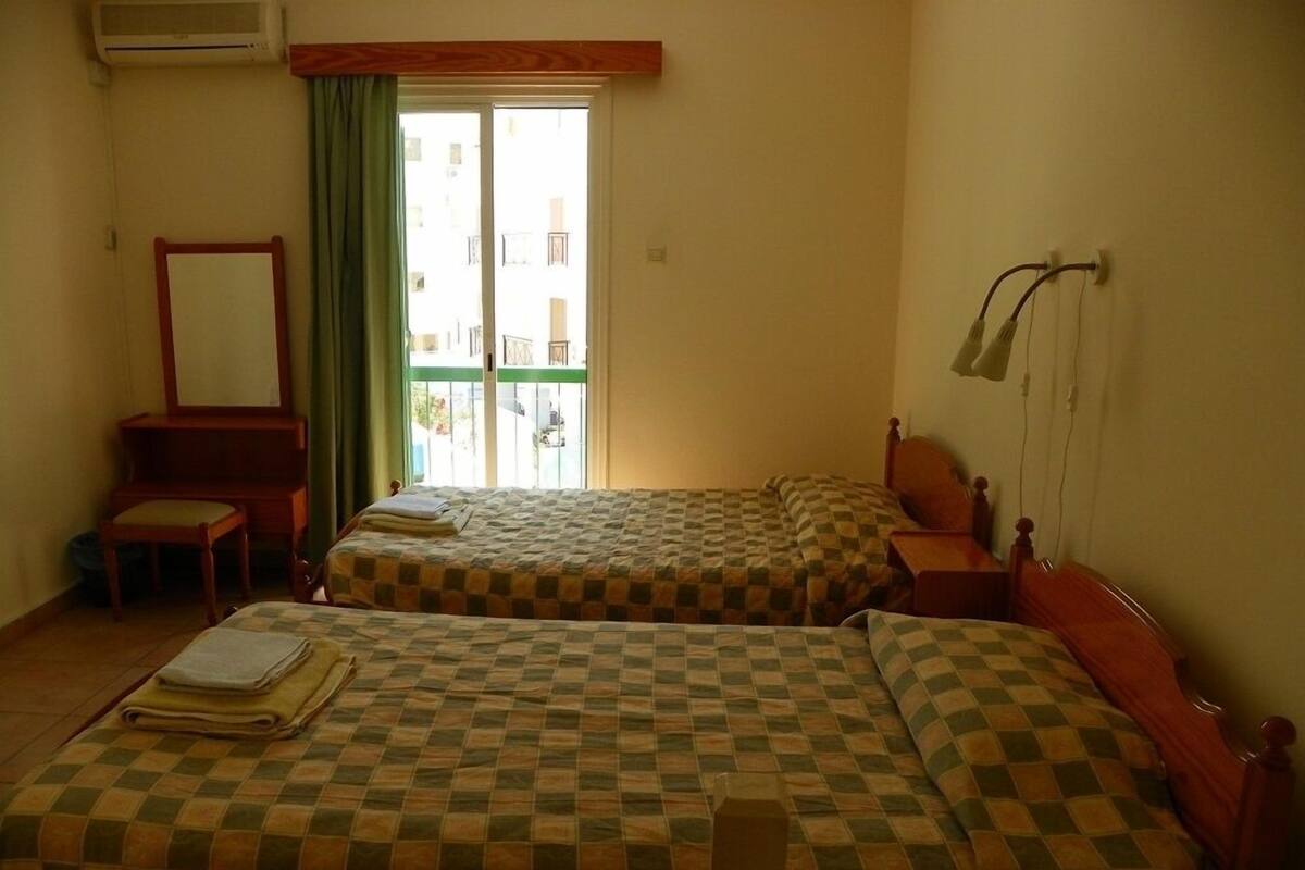 Quarto de hotel com duas camas, uma cadeira de madeira, um espelho e uma janela com vista para edifícios.