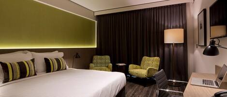 Quarto de hotel com uma cama grande, duas poltronas, uma escrivaninha com um laptop e uma luminária de mesa.