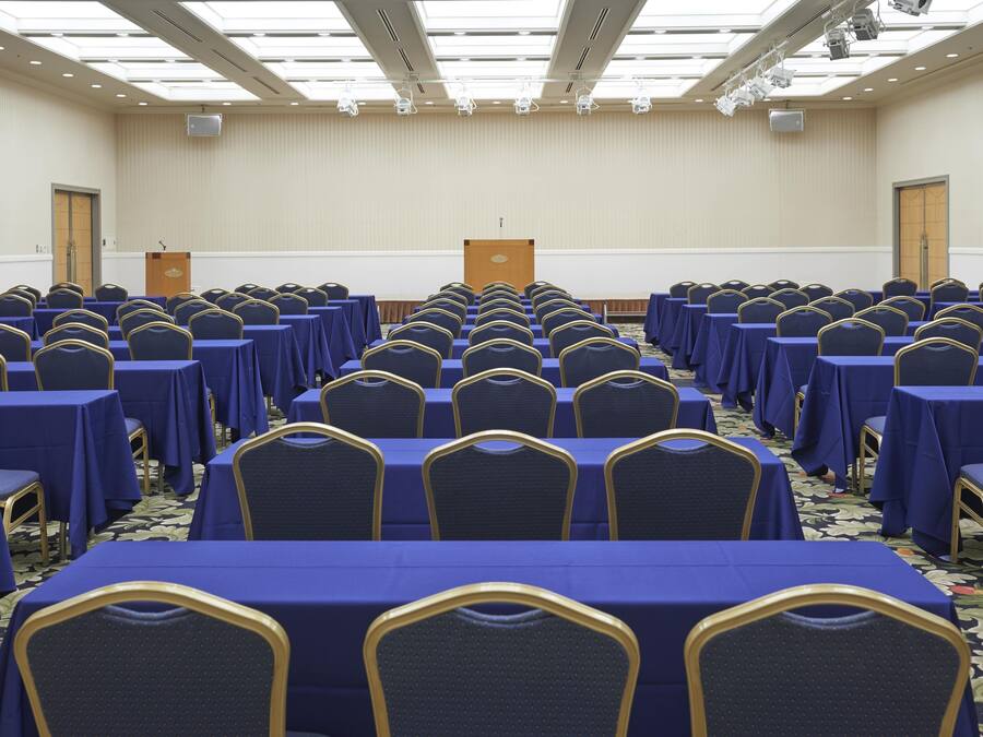 Sala de conferência com fileiras de mesas e cadeiras, um púlpito e uma tela de projeção.