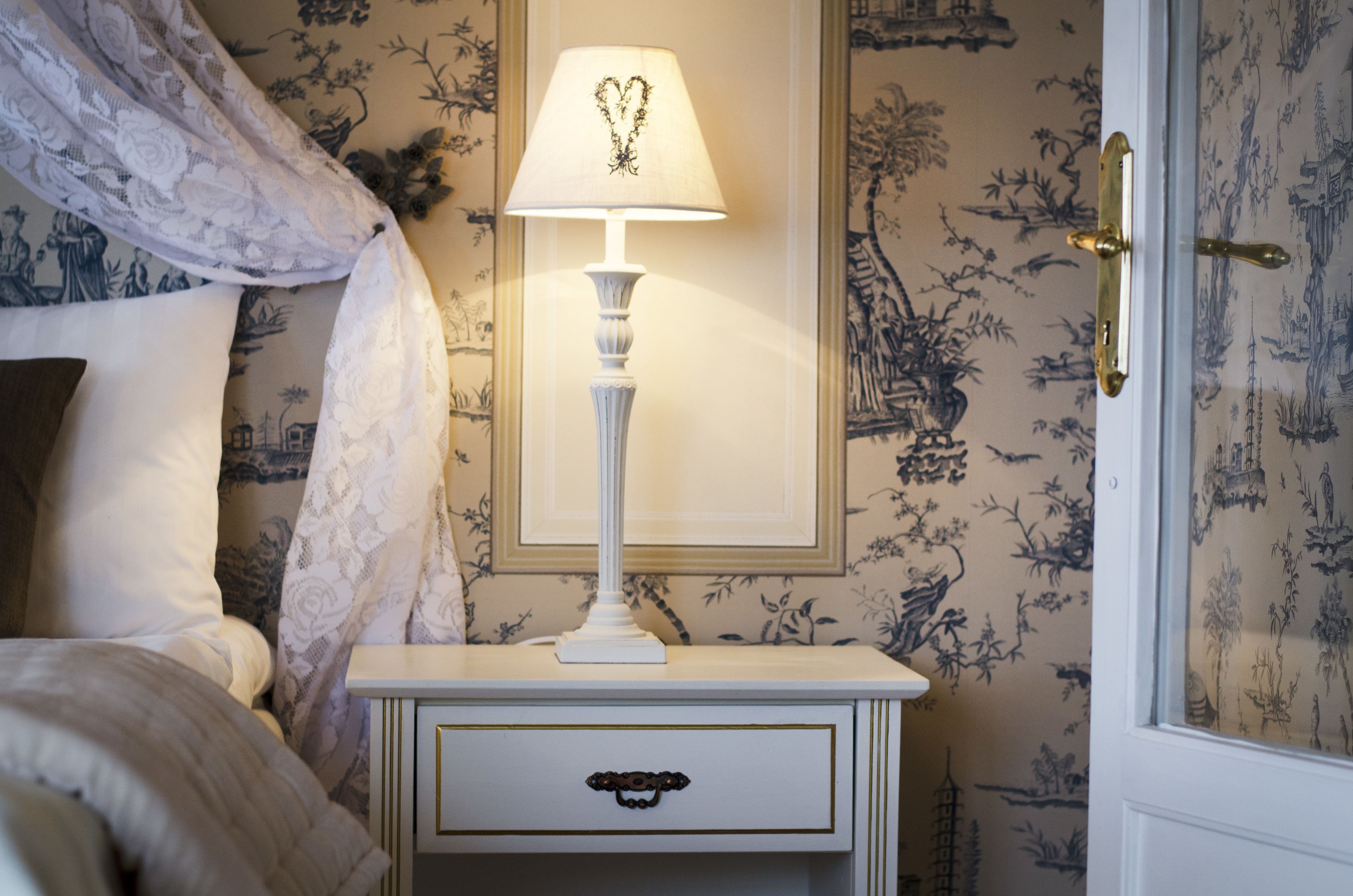 Cama bem arrumada com edredom branco e travesseiro decorativo. Uma mesa de cabeceira branca com abajur e uma gaveta pequena fica ao lado da cama. O papel de parede da parede tem motivos florais.