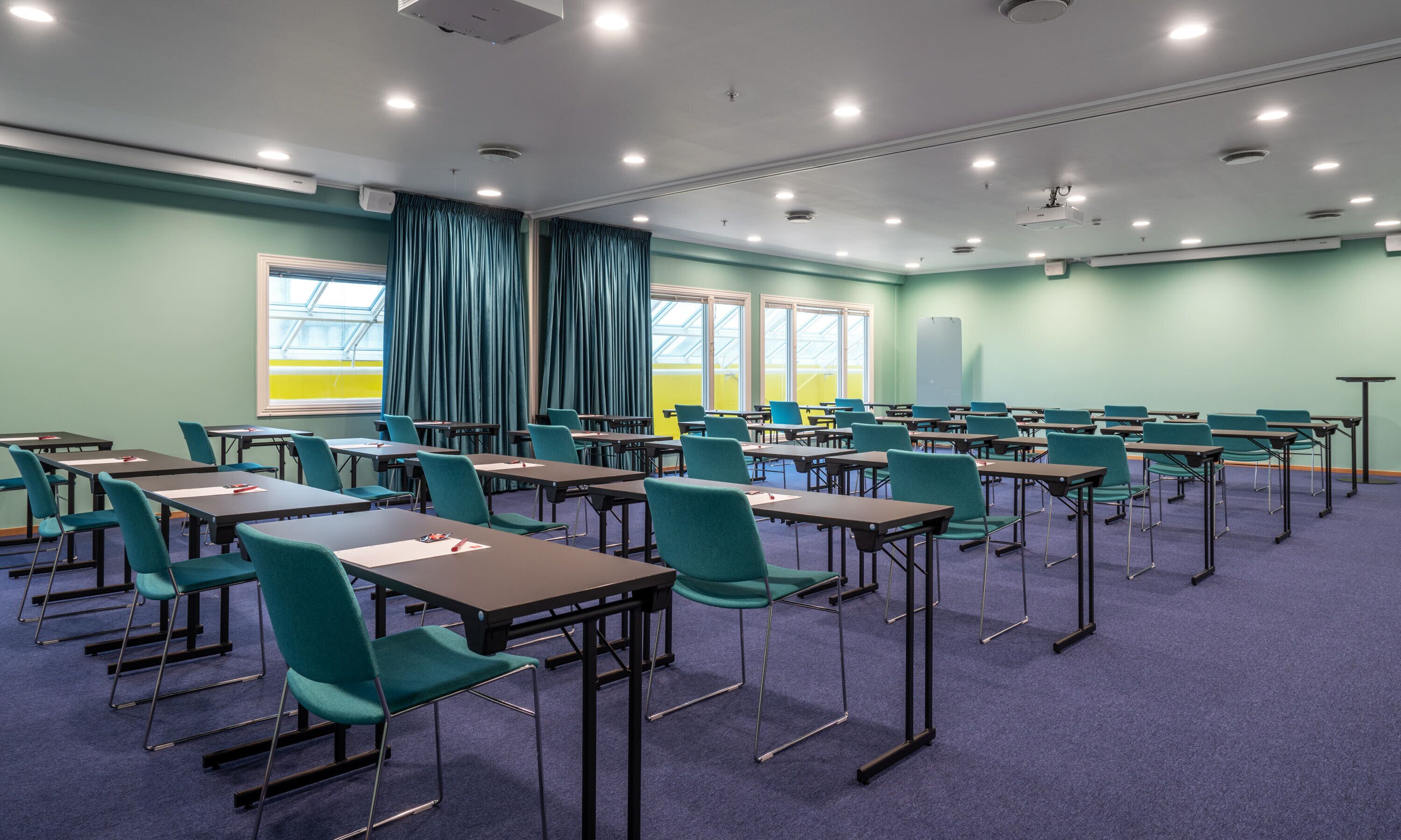 Uma sala de conferências com fileiras de mesas e cadeiras, janelas amplas com cortinas e uma tela de projeção.